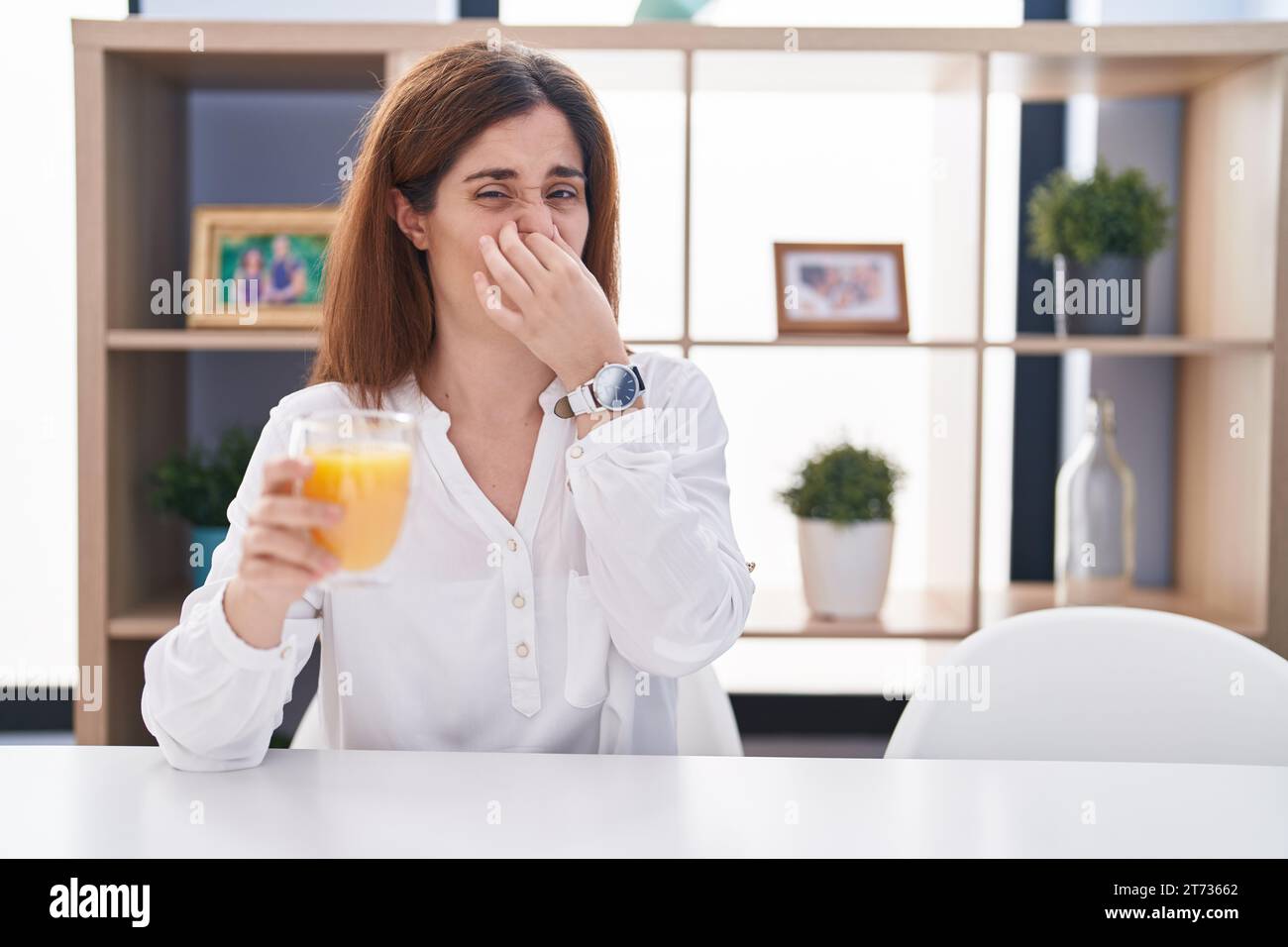 Brunette woman drinking glass of orange juice smelling something stinky ...