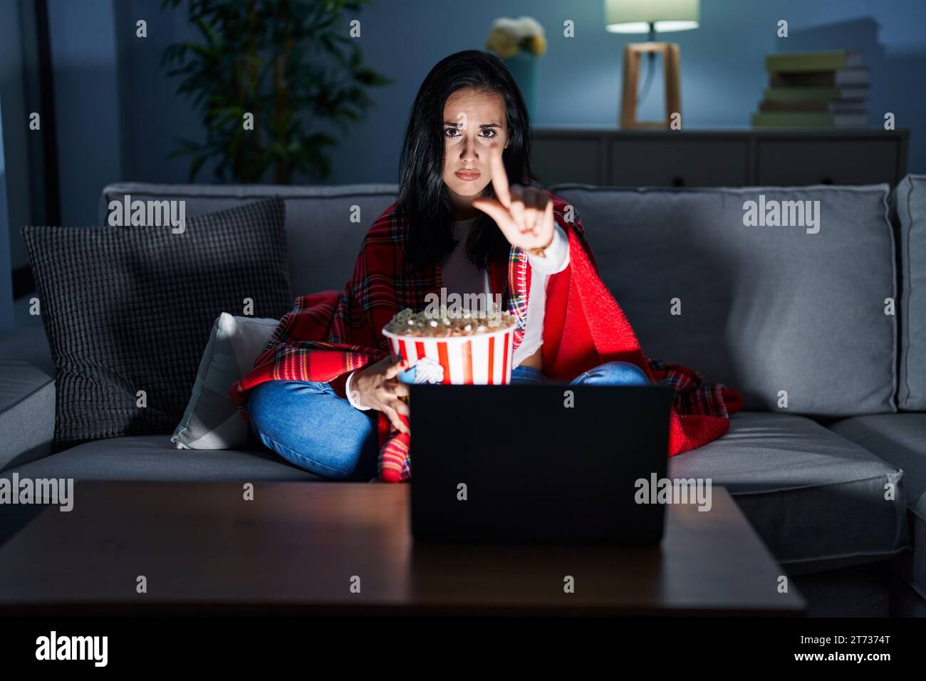 Hispanic woman eating popcorn watching a movie on the sofa pointing ...