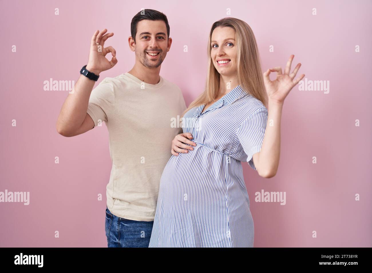 Young couple expecting a baby standing over pink background smiling ...