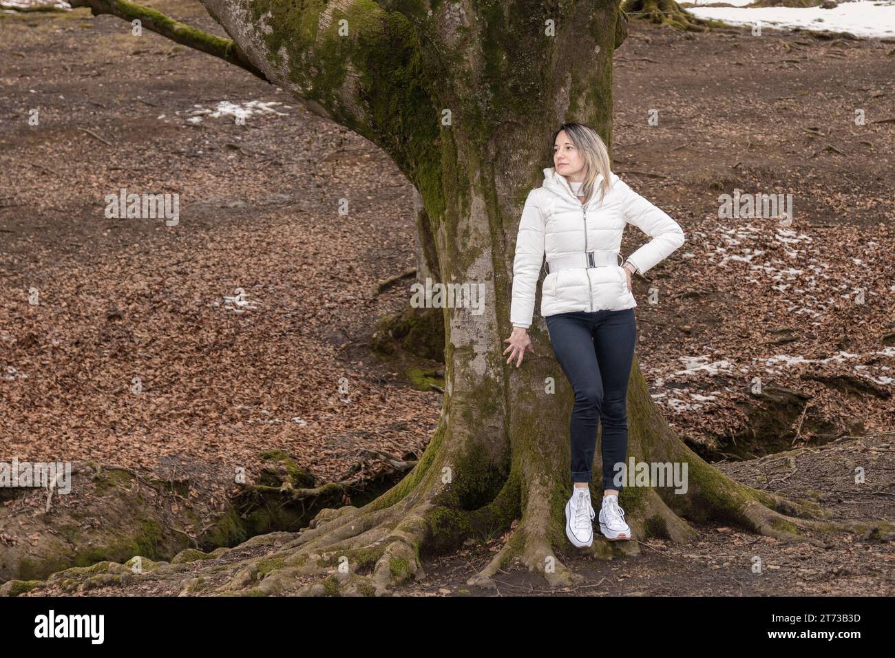 leaning against a tree in a snowy park, wearing a white jacket, blue ...