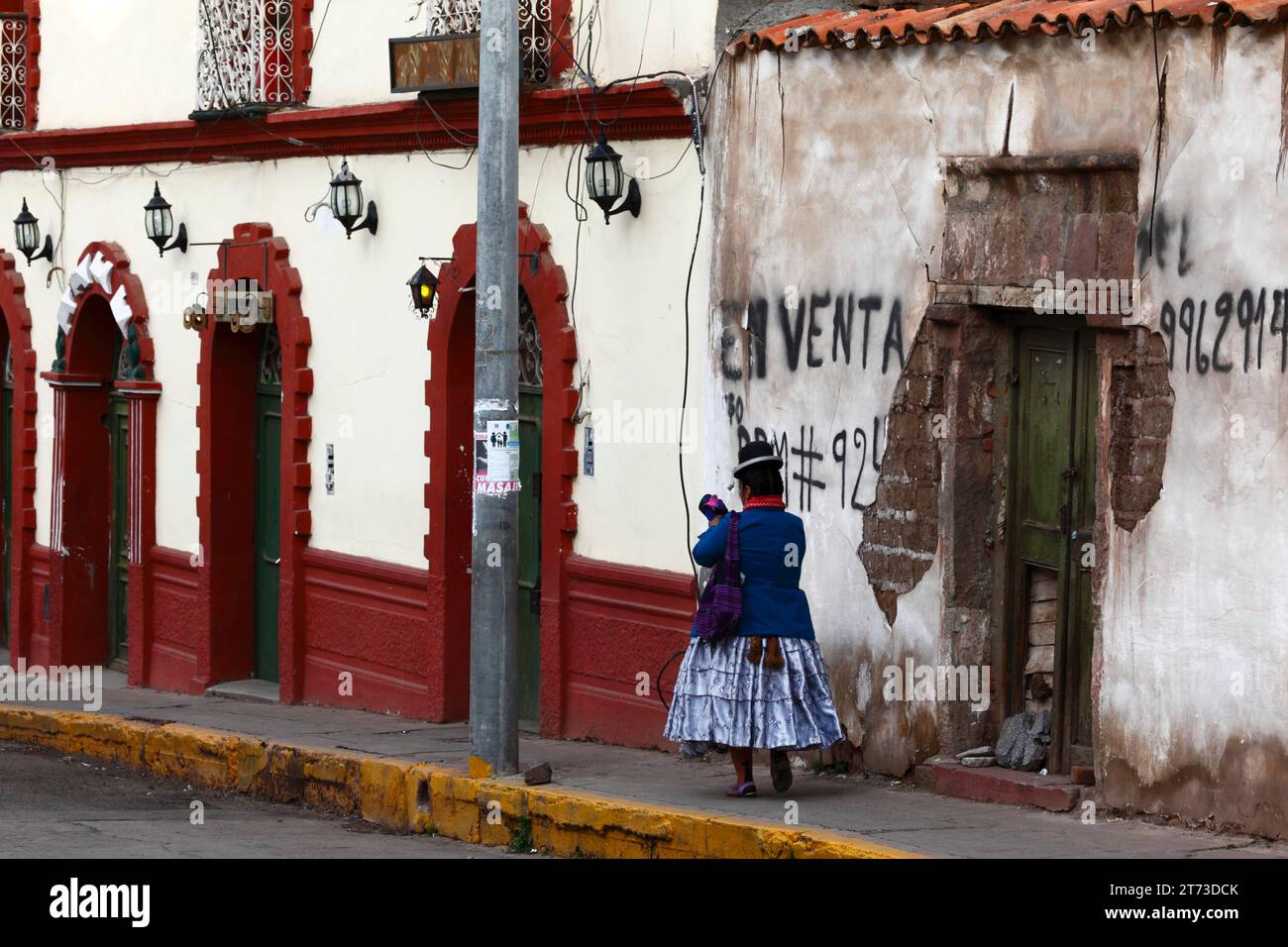 An Aymara woman wearing traditional dress walks past an old house that ...