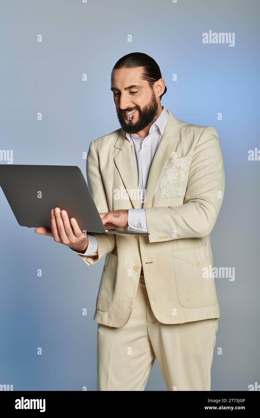 happy bearded arabic man in stylish formal wear using laptop on grey ...