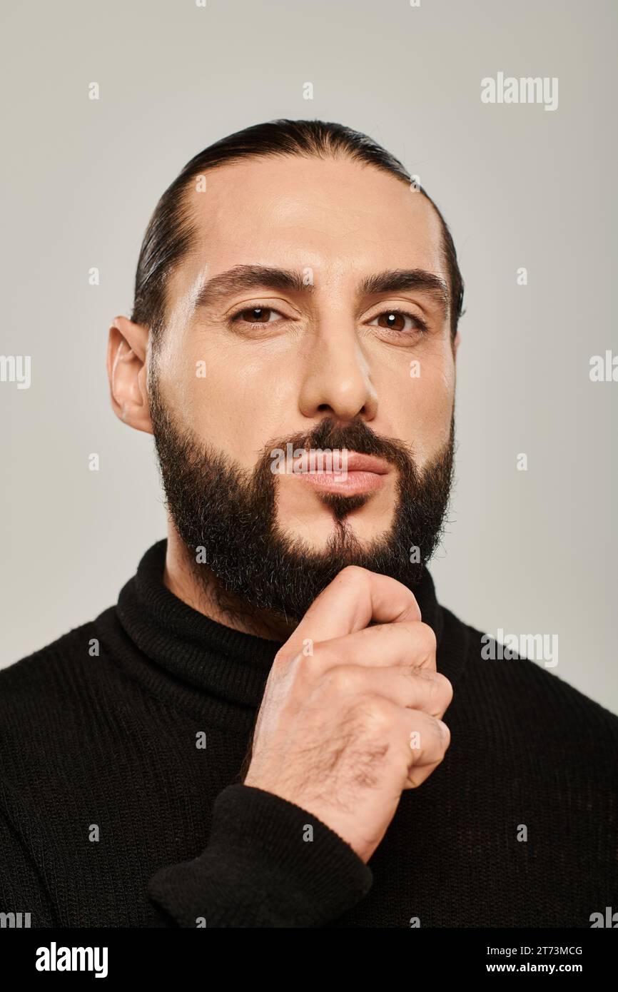 handsome middle eastern man in black turtleneck touching beard on grey ...