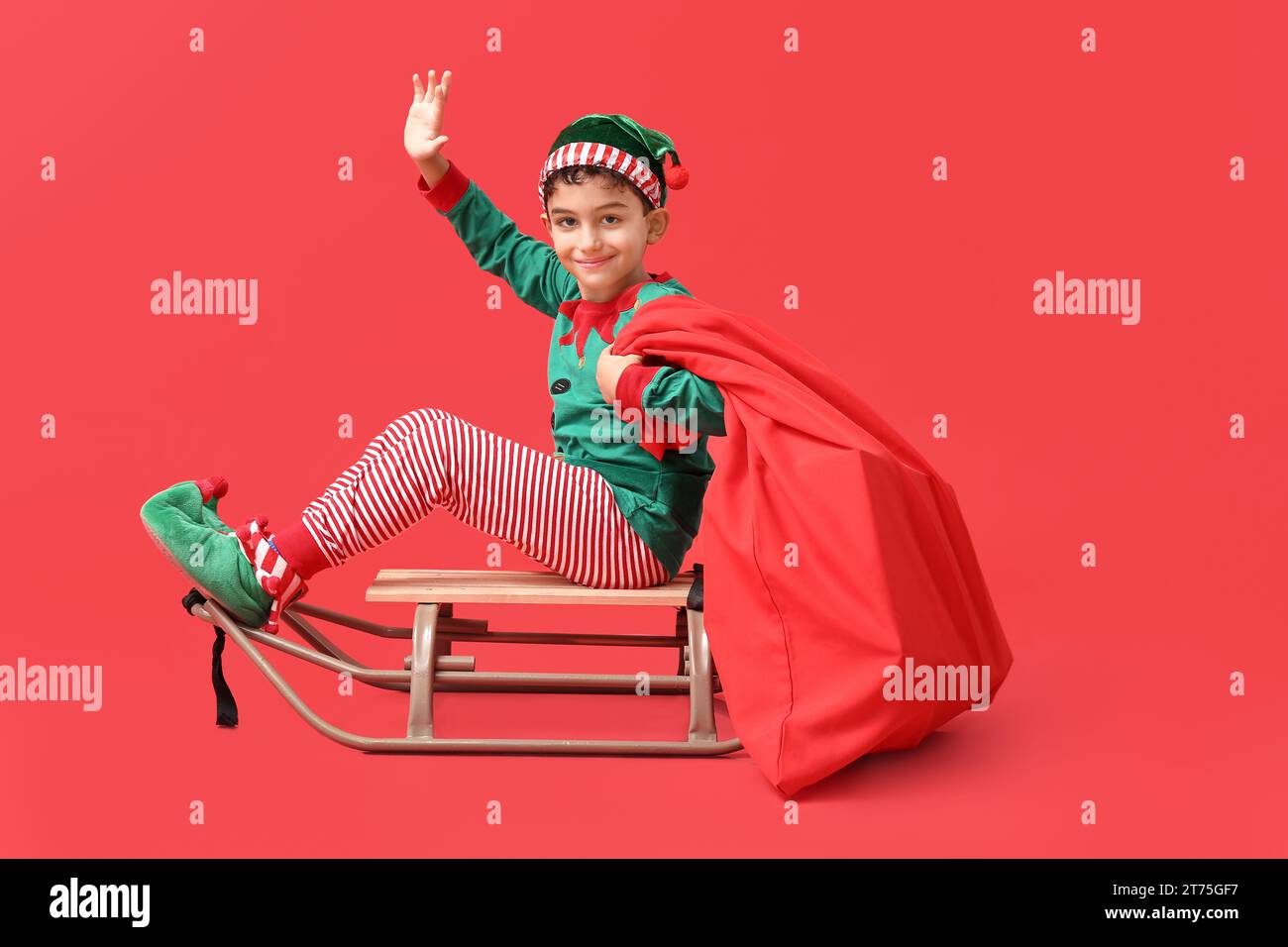 Cute little elf with sled and bag of gifts waving hand on red ...