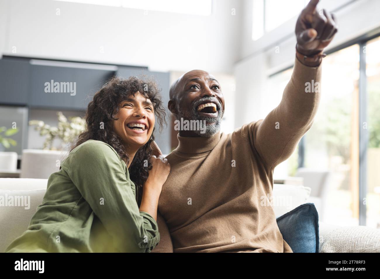 Happy diverse mature couple watching tv pointing and laughing in sunny living room Stock Photo ...
