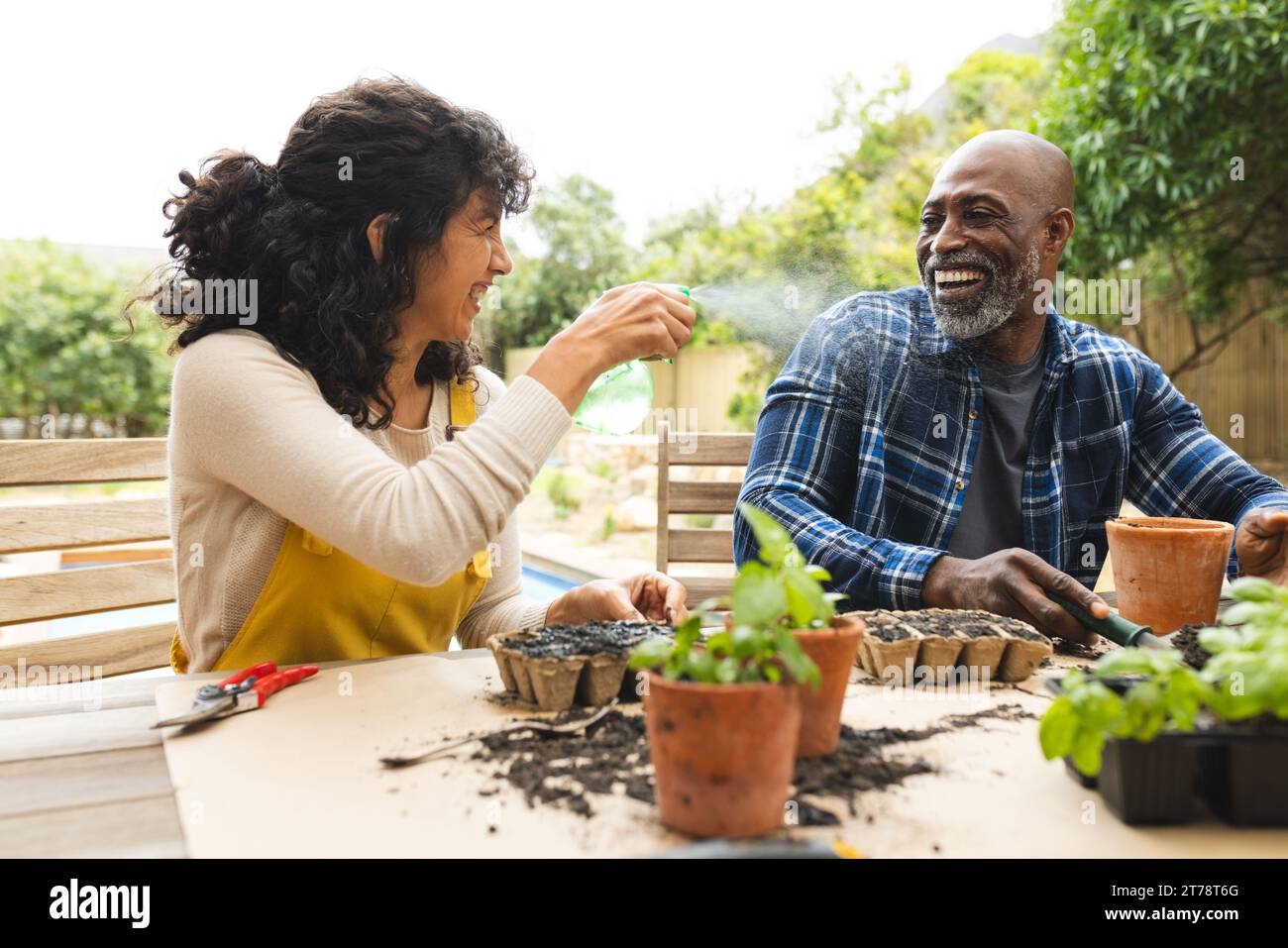 Happy mature diverse couple having fun with water spray, potting plants ...