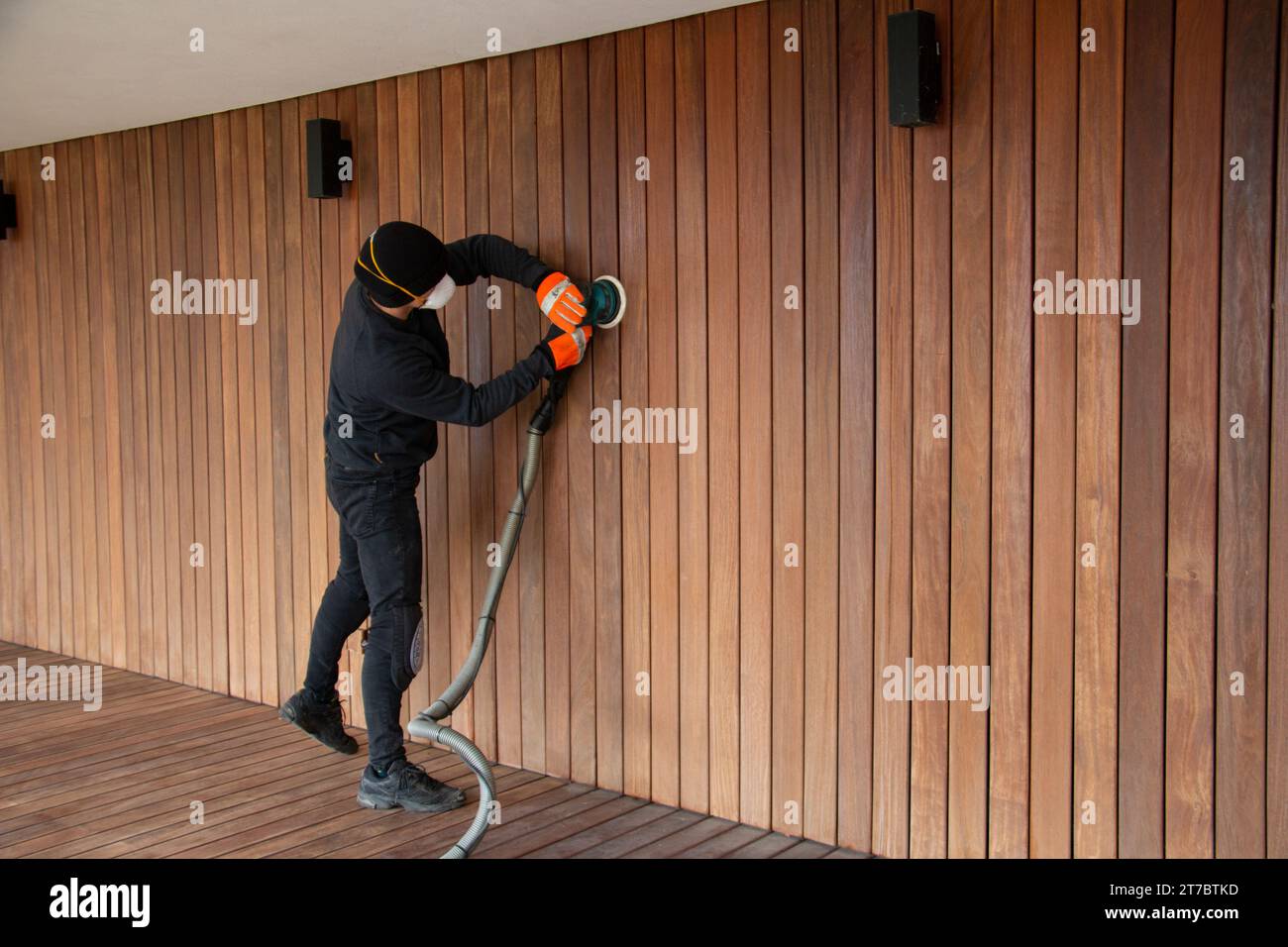 Deck worker in mask sanding hardwood siding with orbital sander, wooden ...