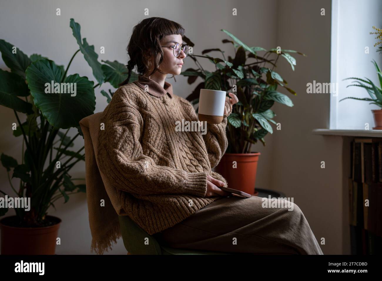 Thoughtful teen girl with cup of tea and smartphone looking at window ...