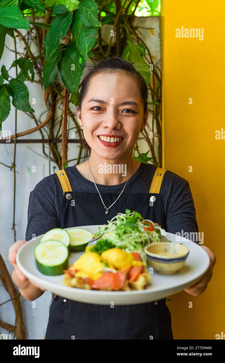 Young vietnamese waitress holding a plate with eggs bennedict waffles ...