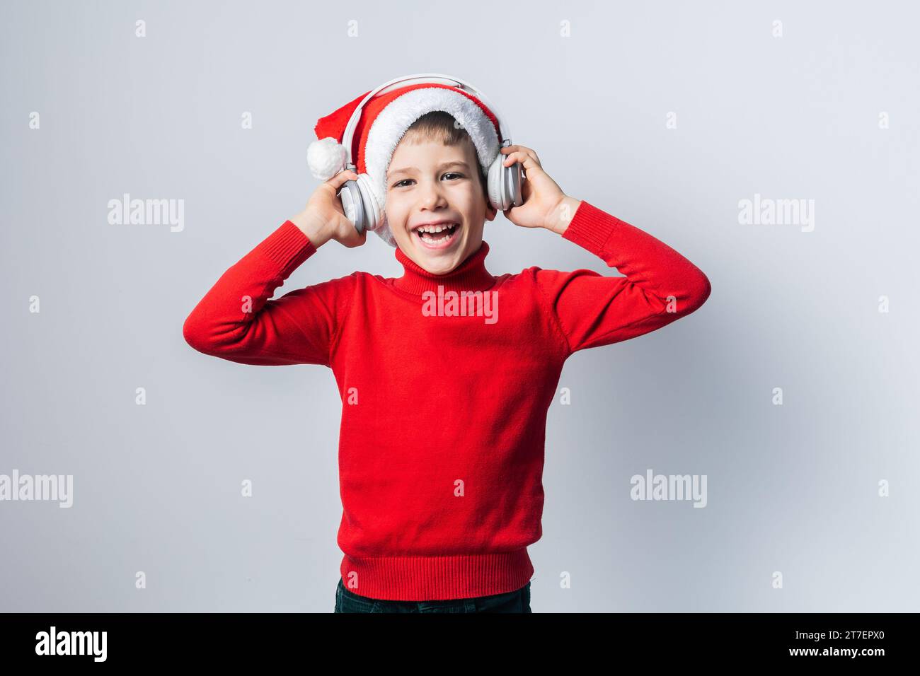 Funny child boy holds listening to Christmas music with Santa Claus hat on white background ...