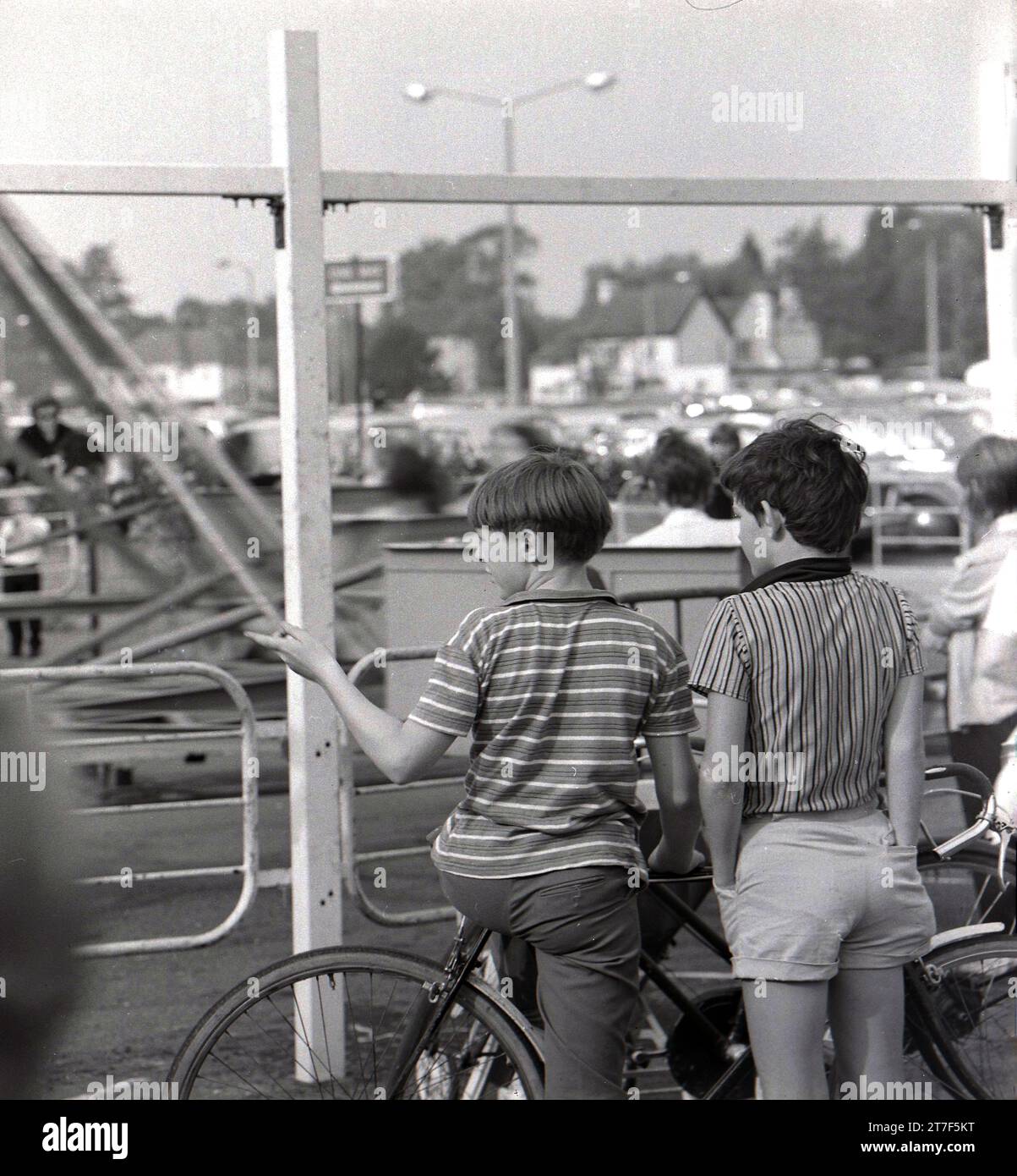 1970s, two young boys with bikes standing outside a barrier watching the activity at a ...