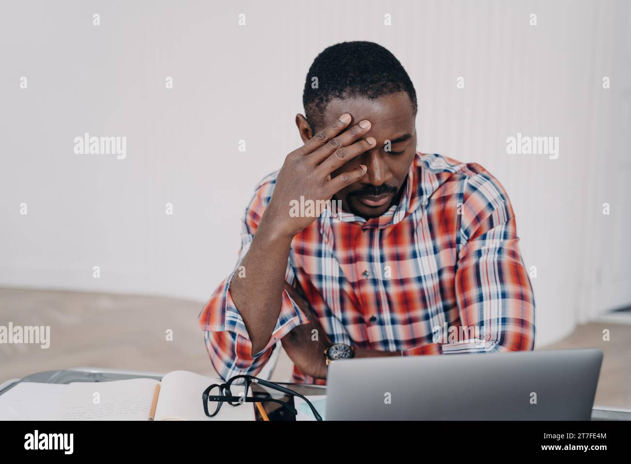 Exhausted Black man with hand on forehead stressing over work at laptop ...