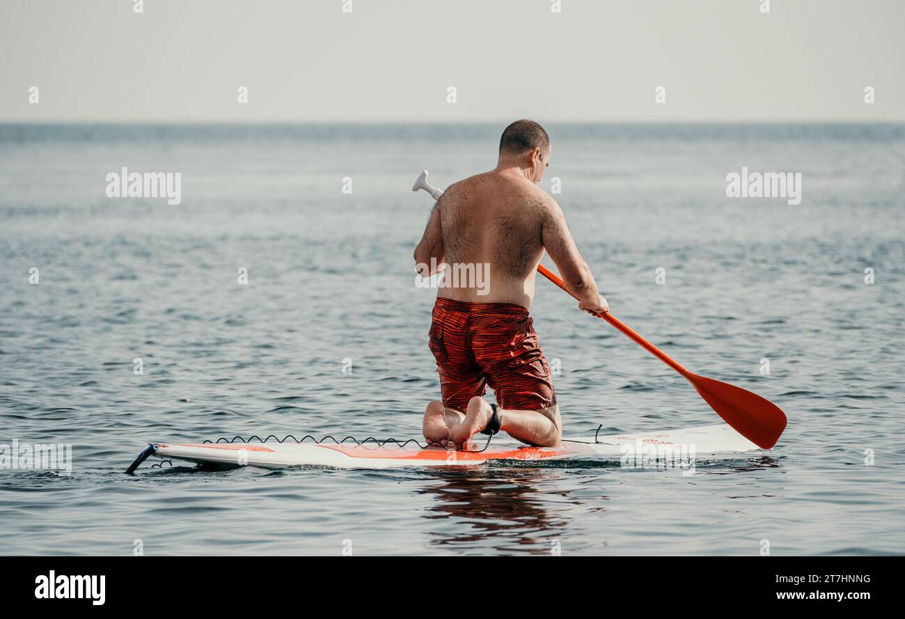 Active mature male paddler with his paddleboard and paddle on a sea at summer. Happy senior man ...