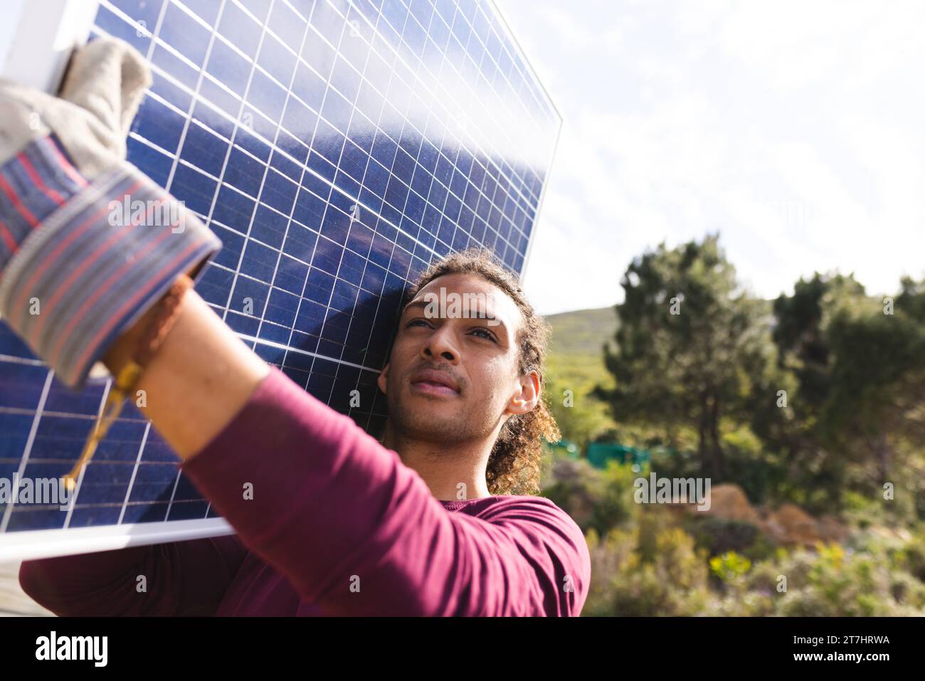 Focused biracial man carrying solar panel and smiling in sunny garden ...