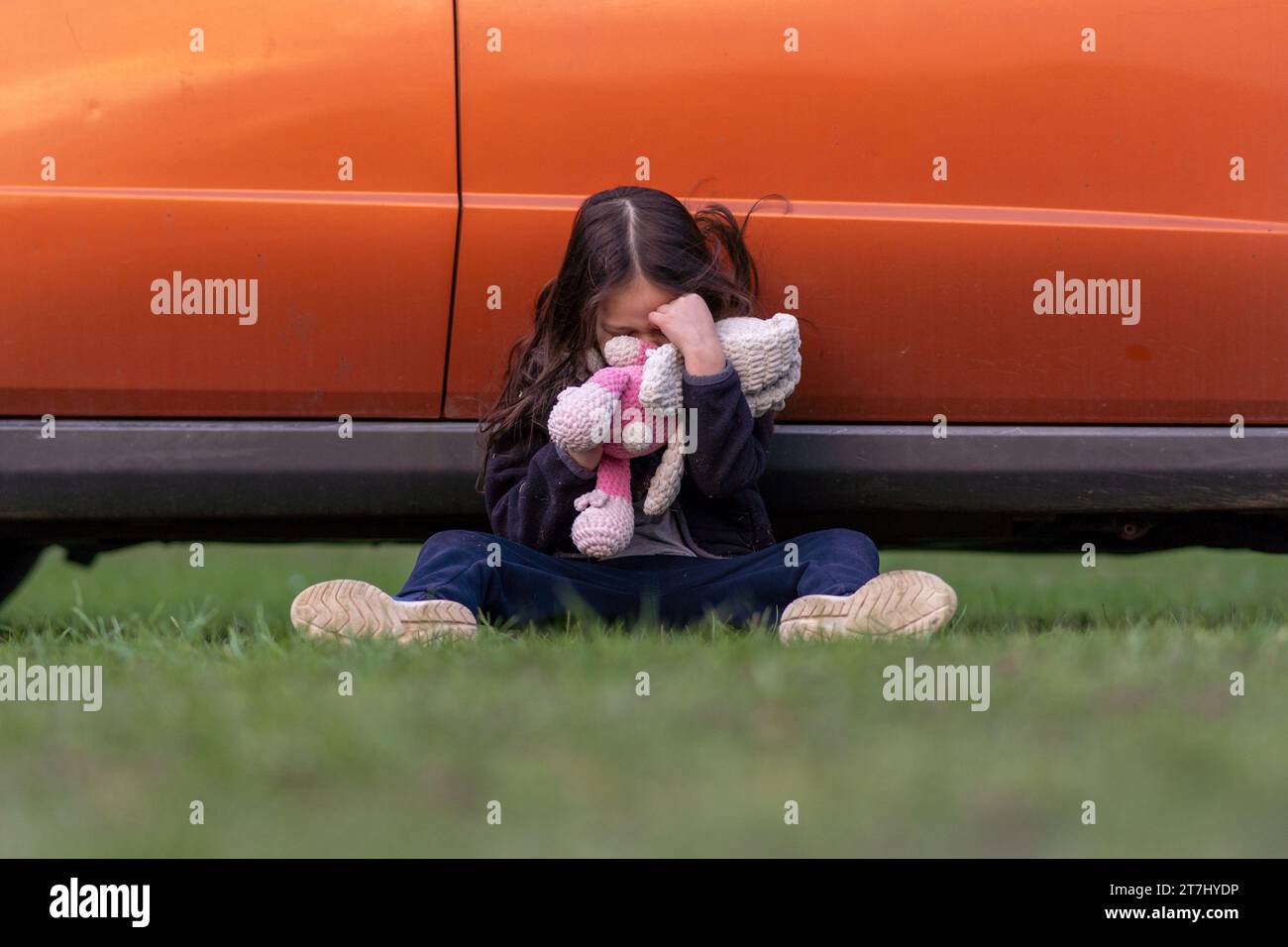 Little lonely girl is crying, hiding her face in toy, sitting on grass ...