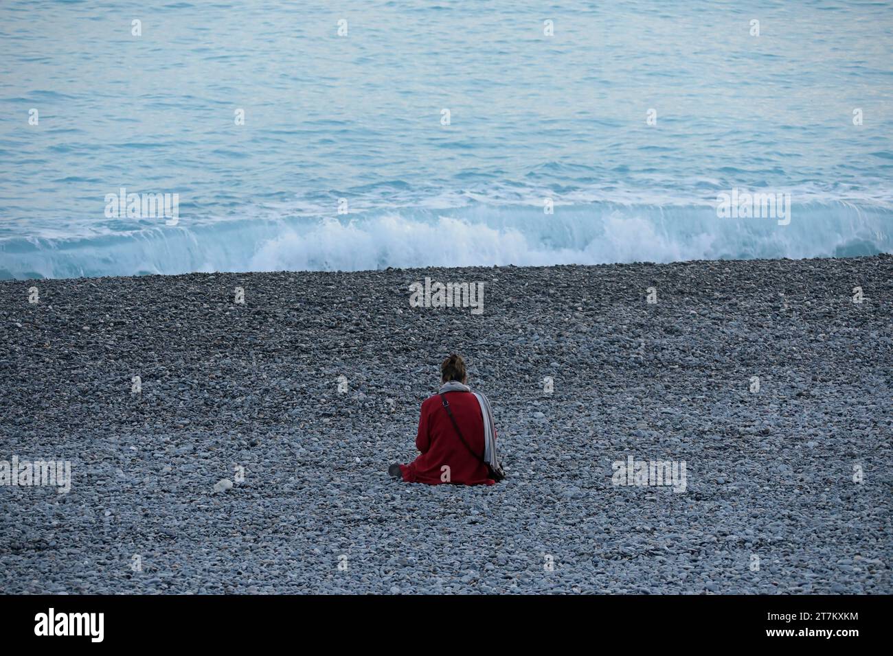 Woman in a red coat sitting alone on the beach at Nice on the French ...
