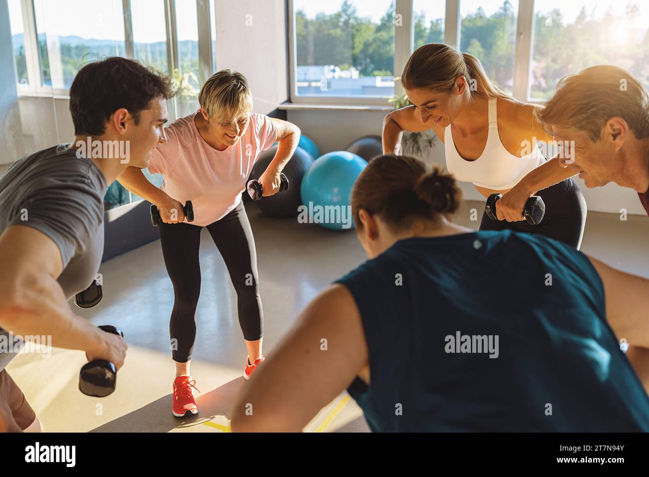 Diverse gym group doing dumbbell exercises, sunlight streaming through ...