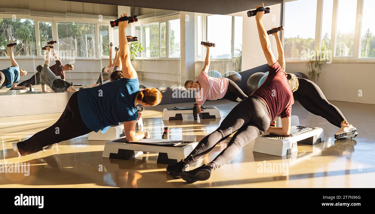 Fitness class performing side plank with dumbbells, sunlit gym, mirror ...