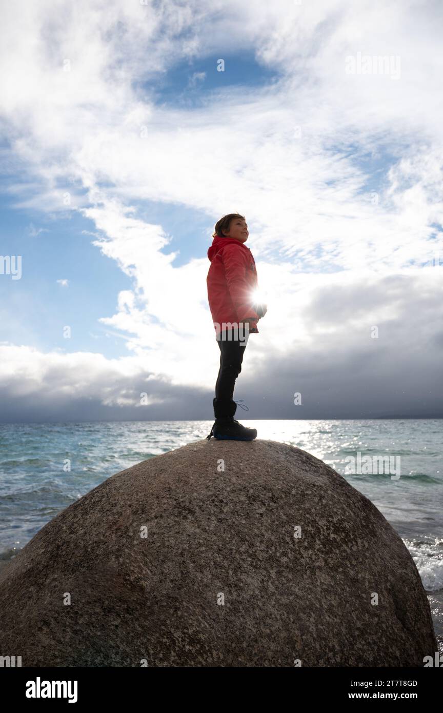 Backlit boy in red jacket stands on top of large boulder with lake ...