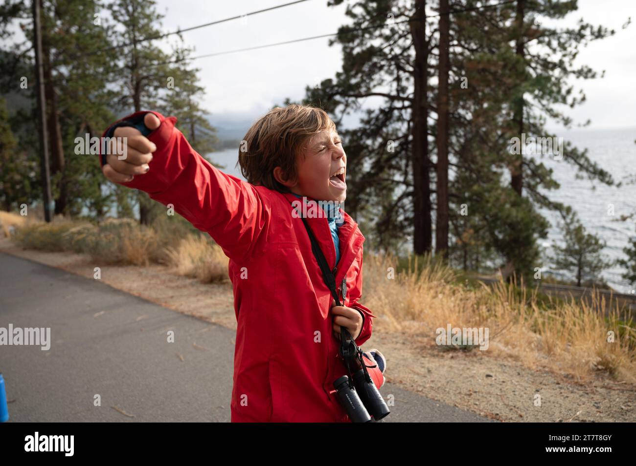Joyful tween in red raincoat with fist lifted on a trail near lake ...