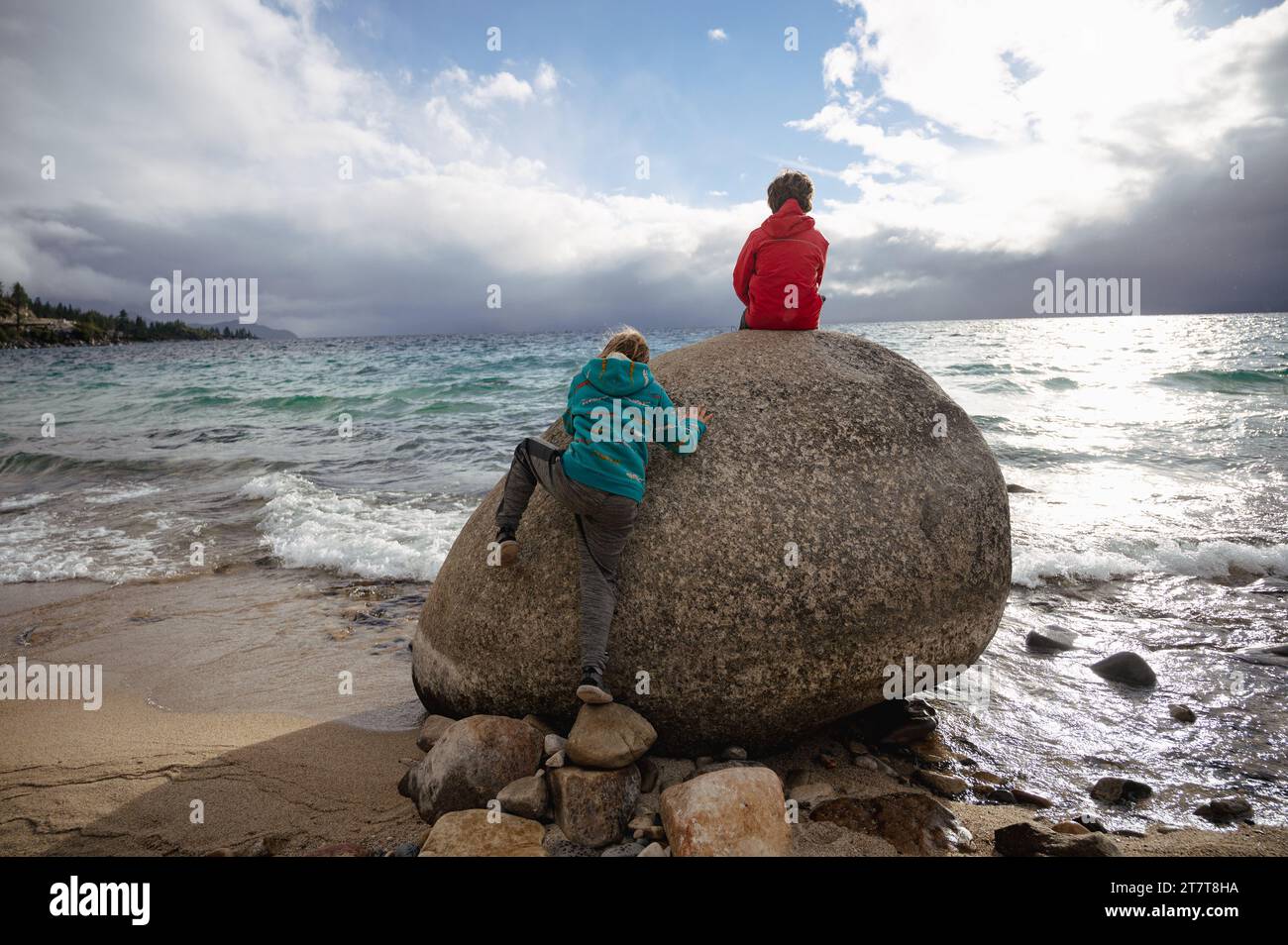 boy sitting on top of boulder at lakeshore as brother climbs up too ...