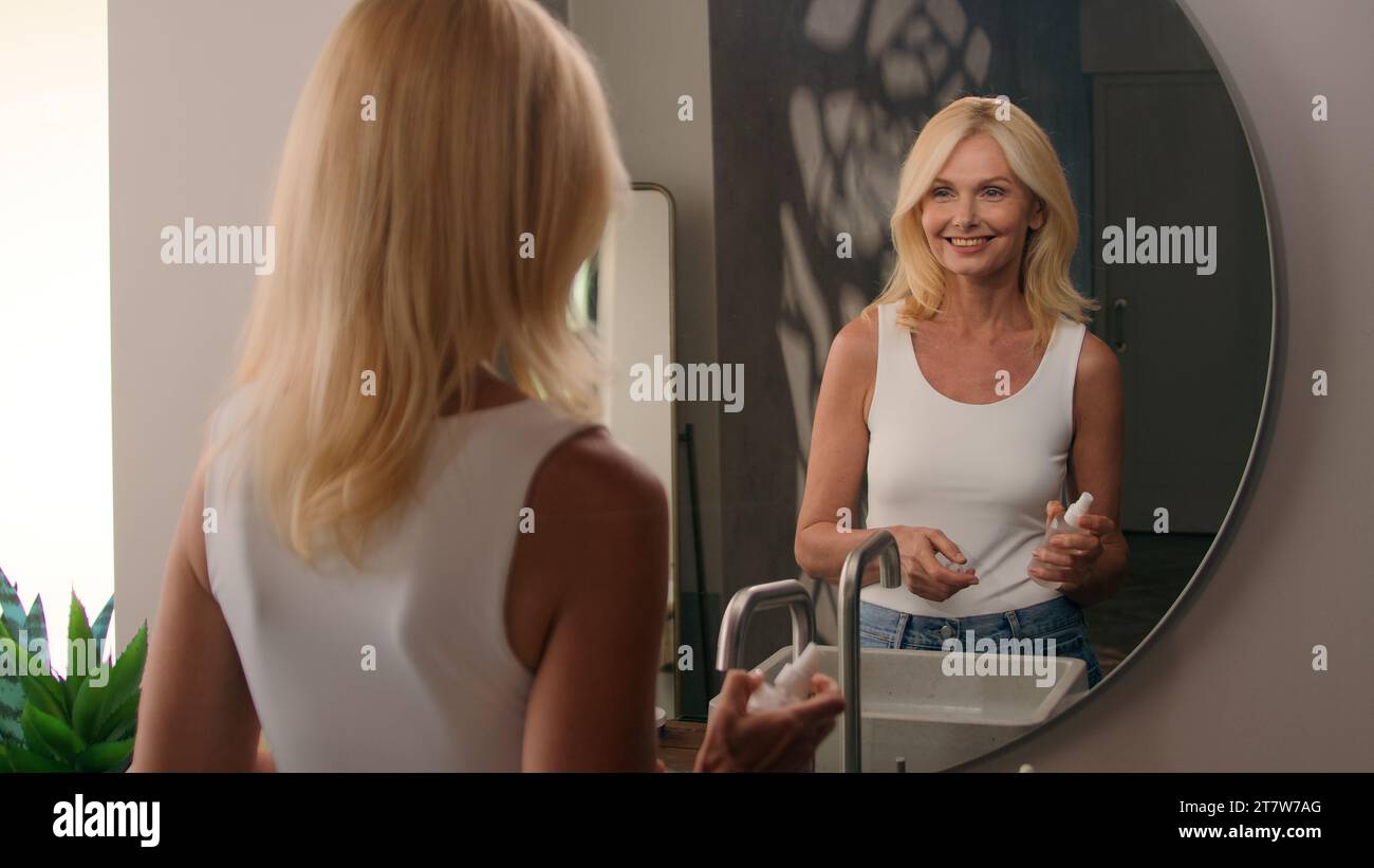 Smiling Caucasian senior mature woman look in bathroom mirror apply ...