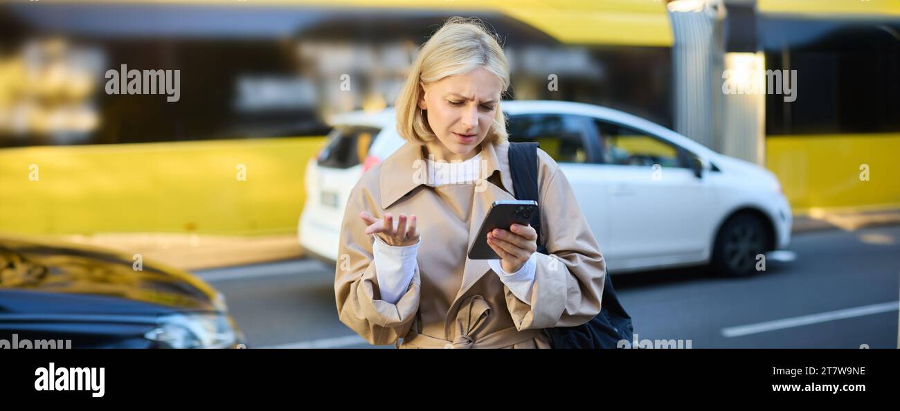 Portrait of young confused blonde woman, standing on busy street with ...