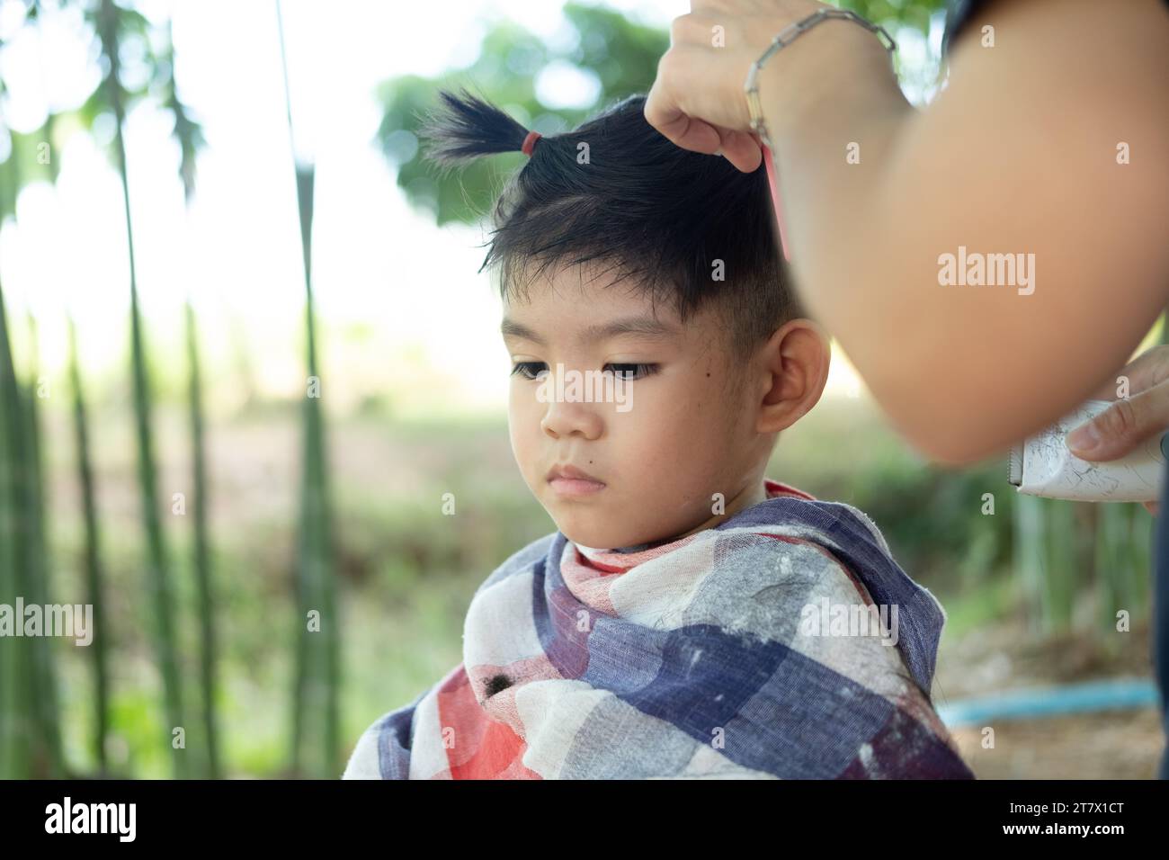 Barber cutting hair of an Asian boy In an open space filled with trees ...