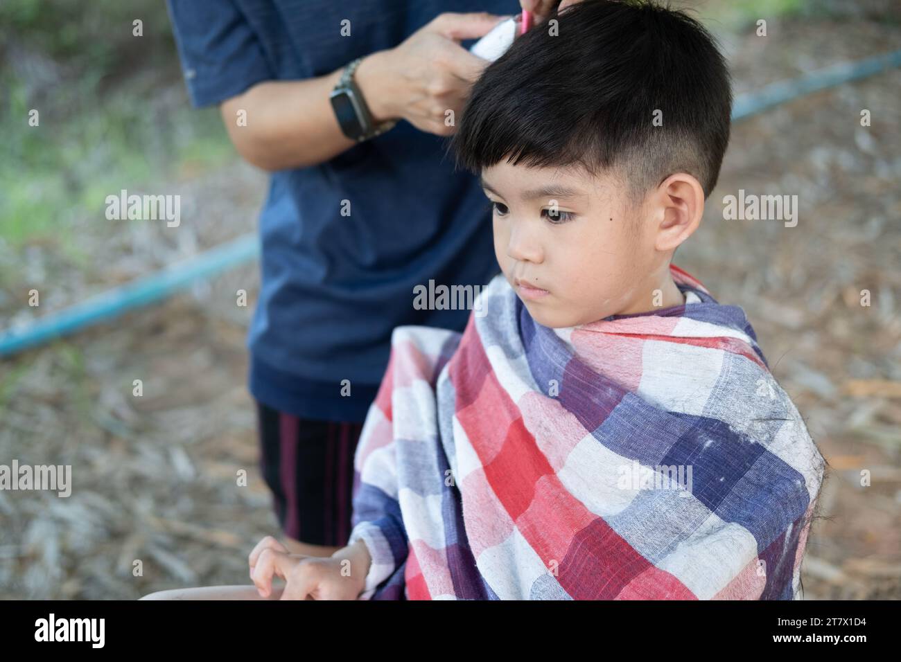 Barber cutting hair of an Asian boy In an open space filled with trees ...