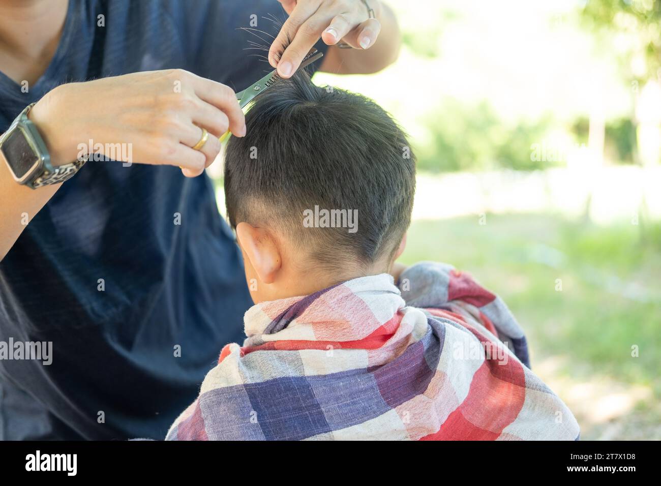 Barber cutting hair of an Asian boy In an open space filled with trees ...