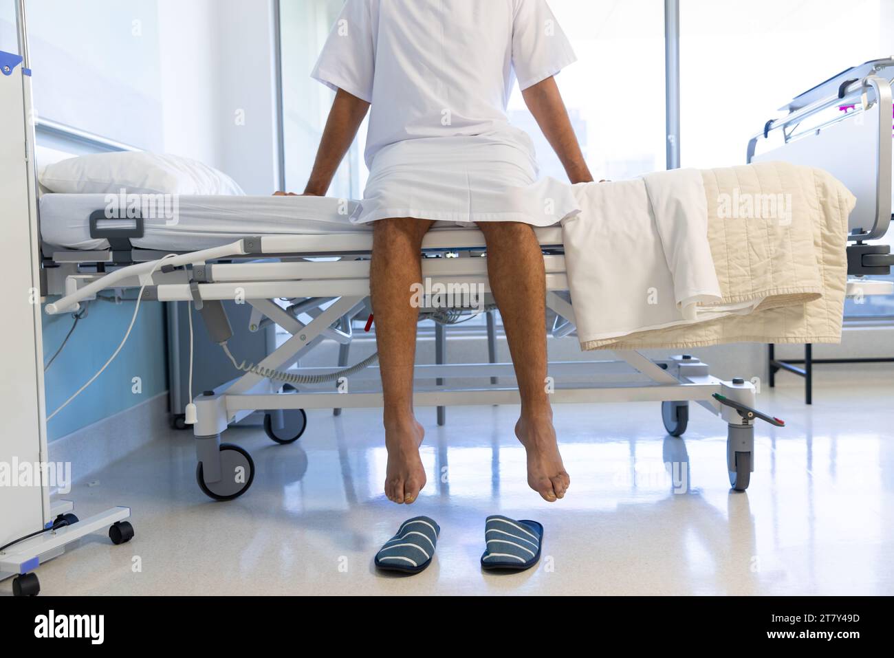 Lowsection of biracial male patient sitting on bed in sunny hospital ...