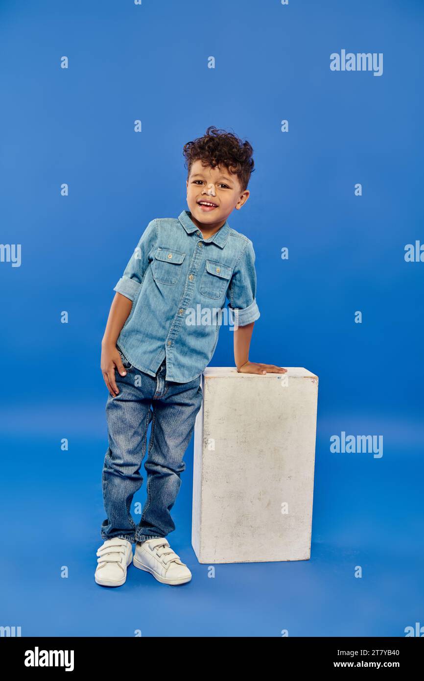 happy and curly african american preschooler boy in denim clothes leaning on white cube on blue ...