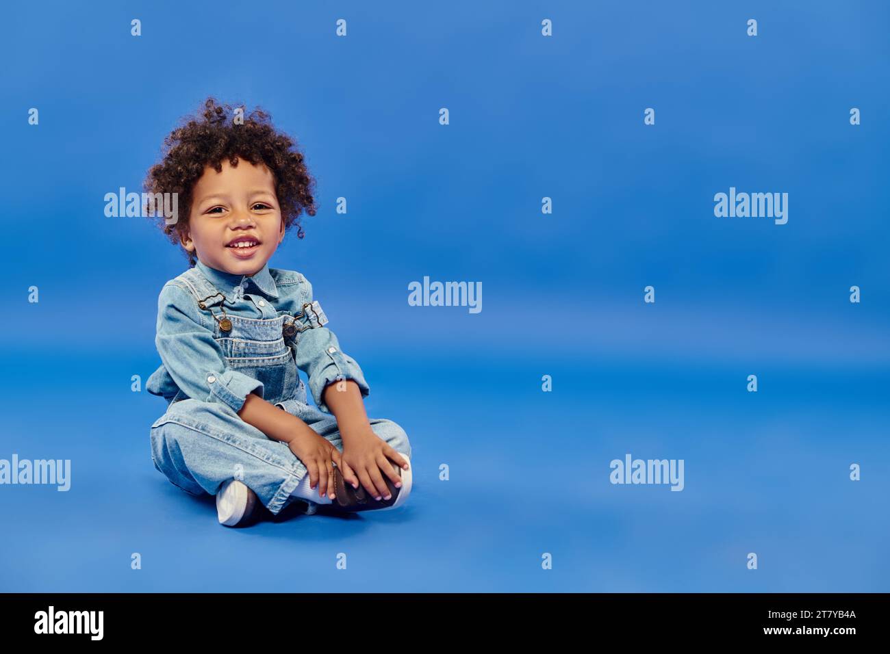 joyful and curly african american toddler boy in denim clothes sitting ...