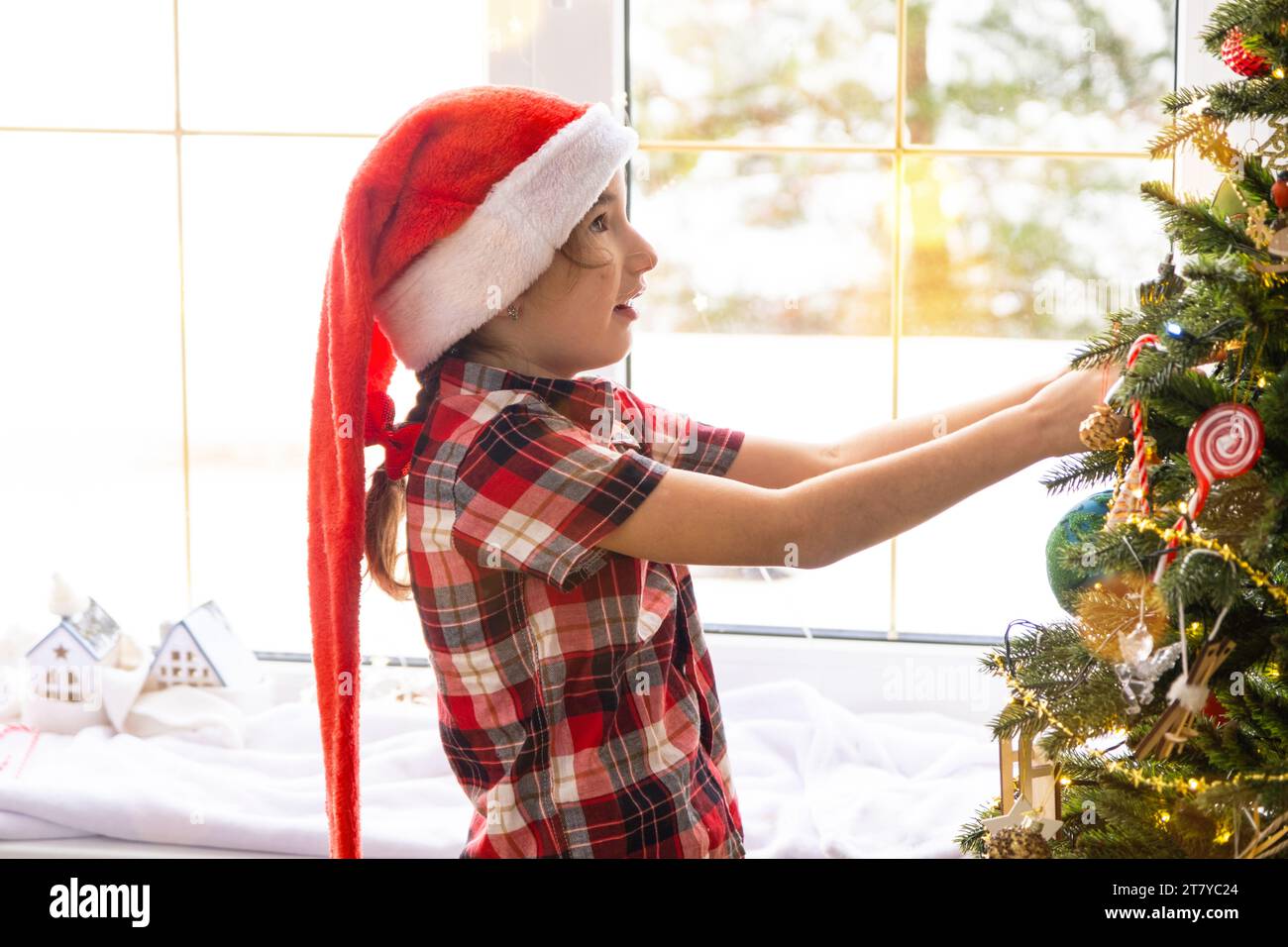 Happy girl in a Santa hat decorates the Christmas tree with colorful ...