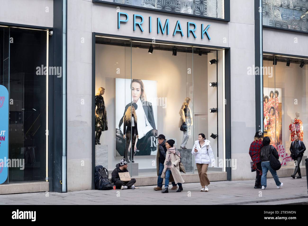 Shoppers and visitors outside walk past a man sitting on the pavement ...
