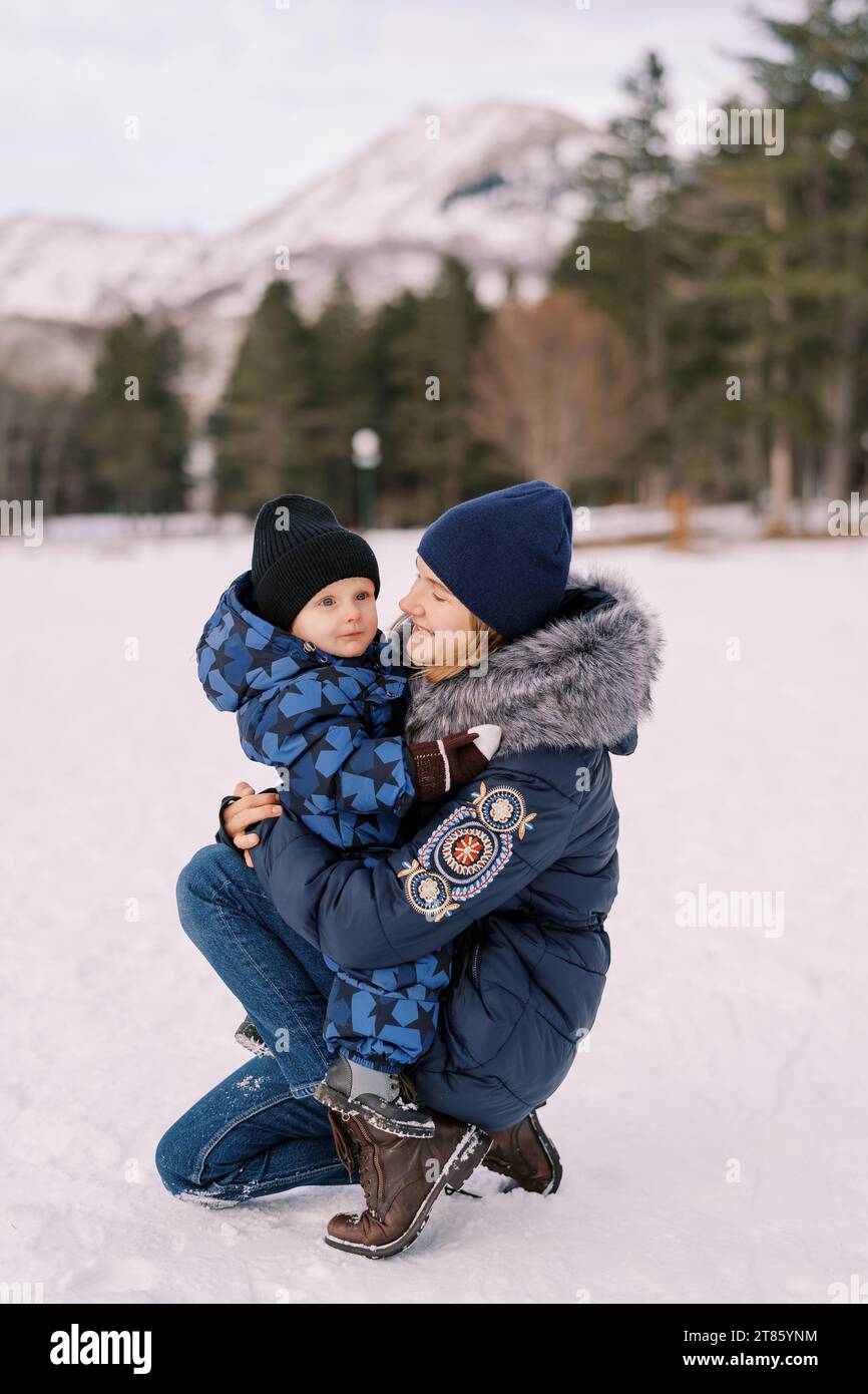 Mom looks at a little boy in her lap while squatting on a snowy lawn ...