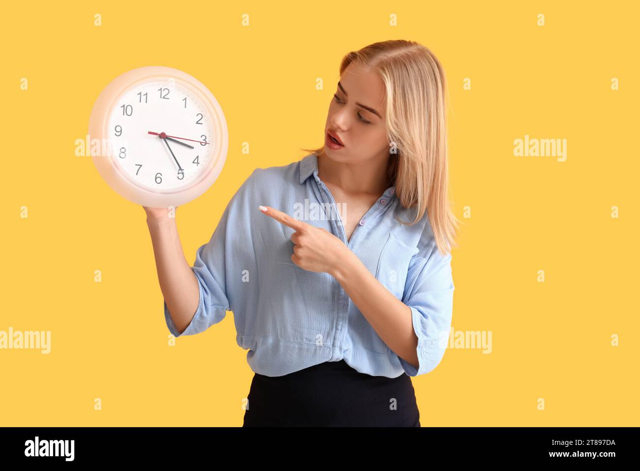 Young businesswoman pointing at clock on yellow background. Time ...