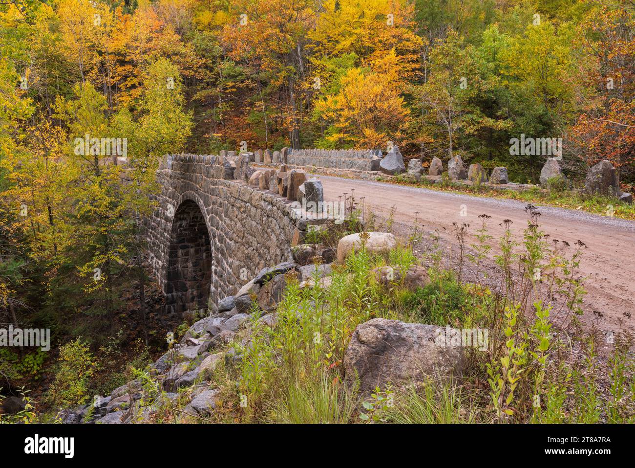 A gravel road crossing a stone arch bridge in the woods during autumn ...