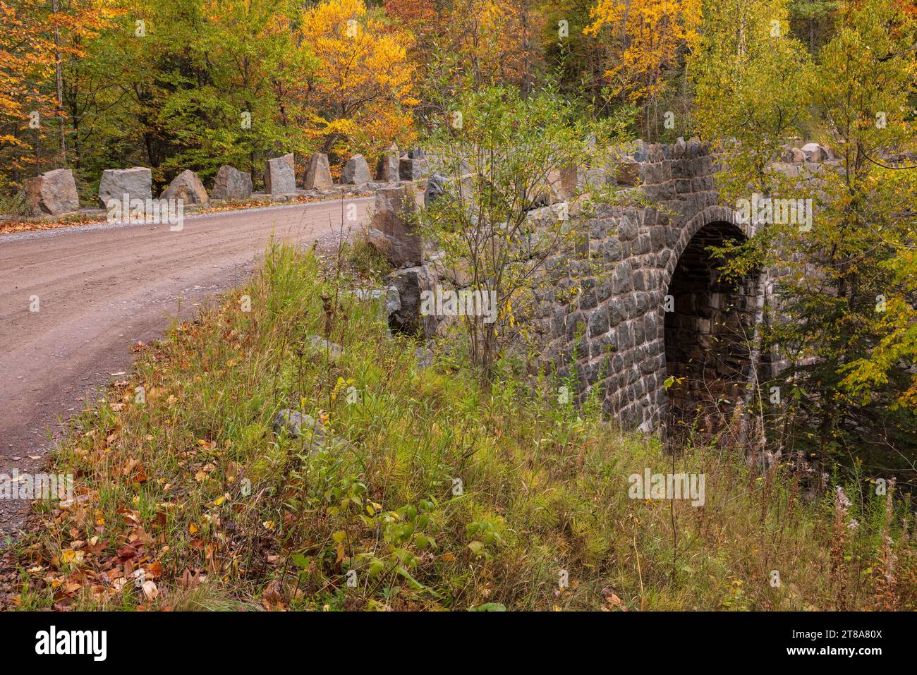 A gravel road crossing a stone arch bridge in the woods during autumn ...