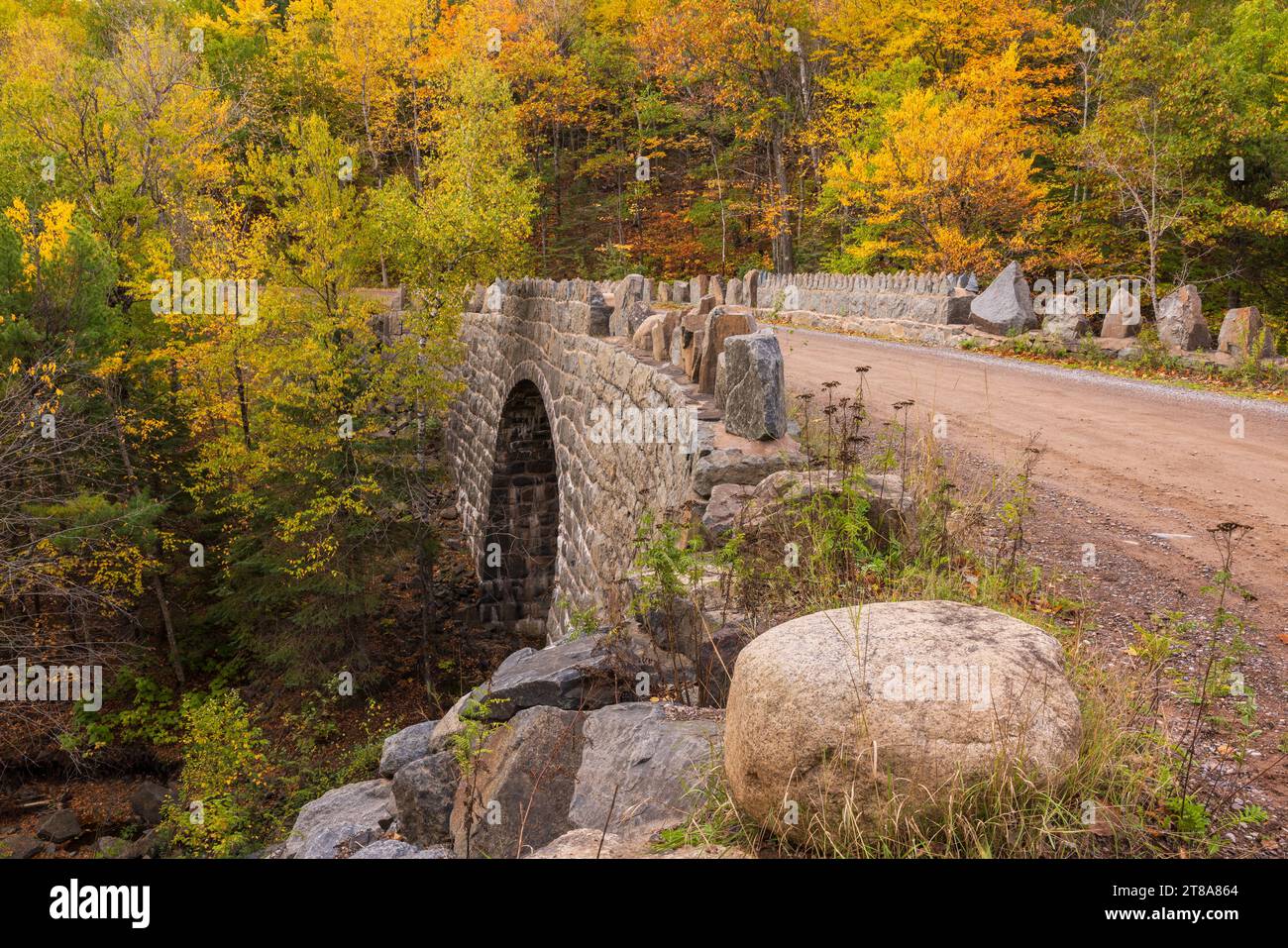 A gravel road crossing a stone arch bridge in the woods during autumn ...