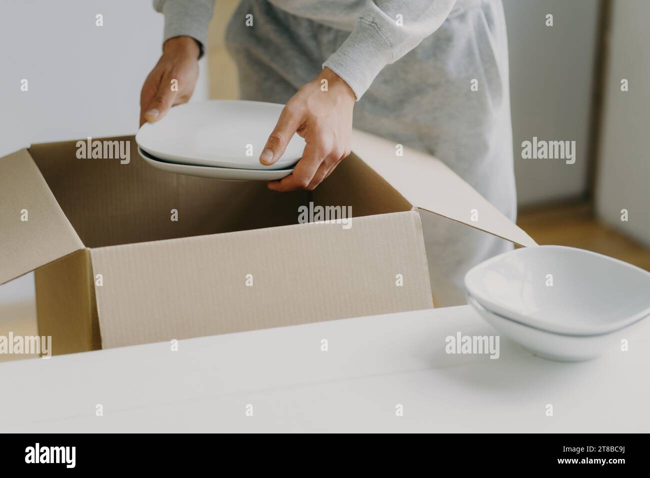 Hands carefully unpacking white dishes from a cardboard box on a table ...