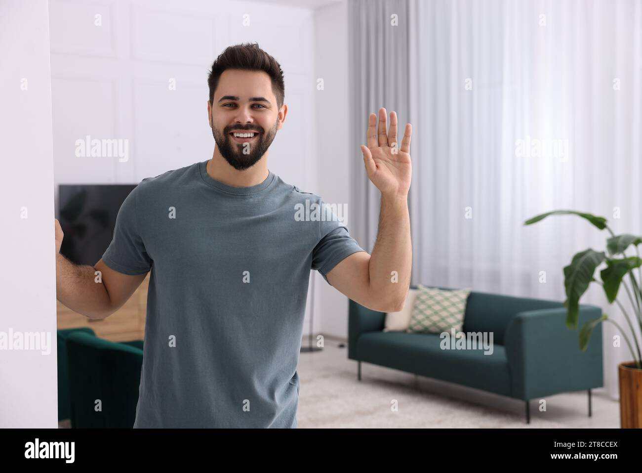 Happy man waving near white wall at home, space for text. Invitation to ...