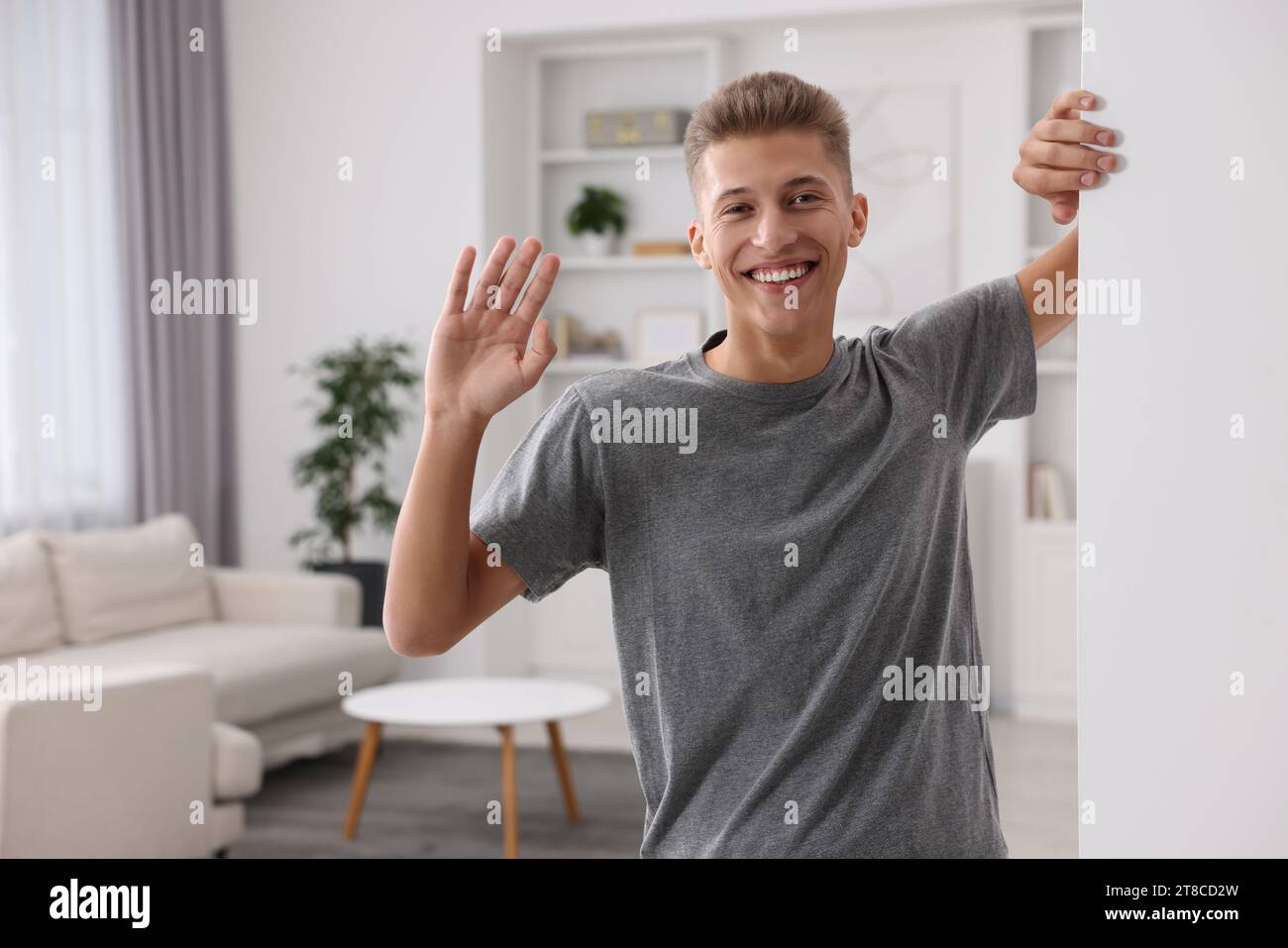 Happy man waving near white wall at home, space for text. Invitation to ...
