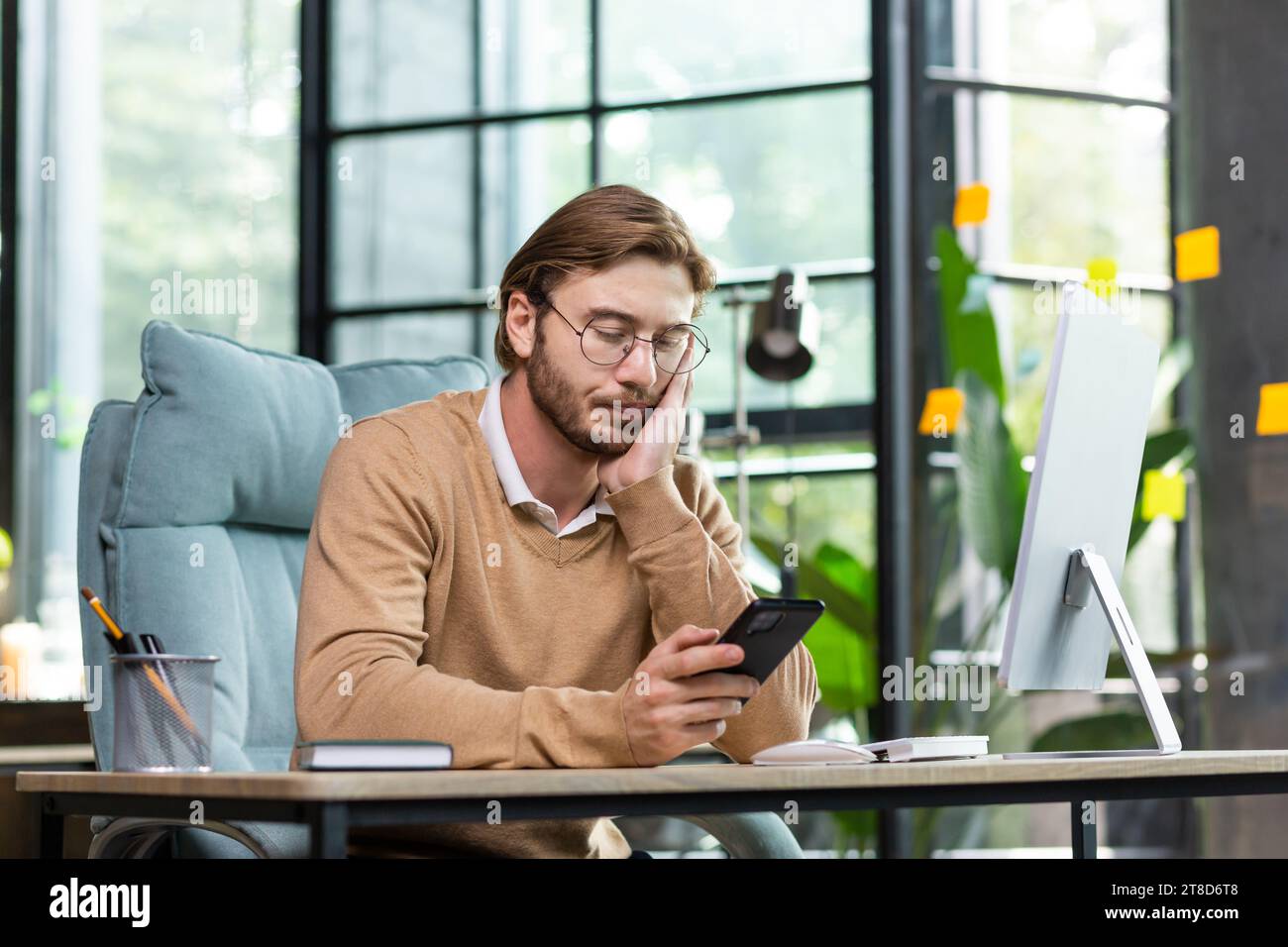 A tired man sits at the table with a phone in his hands, looks sadly at ...