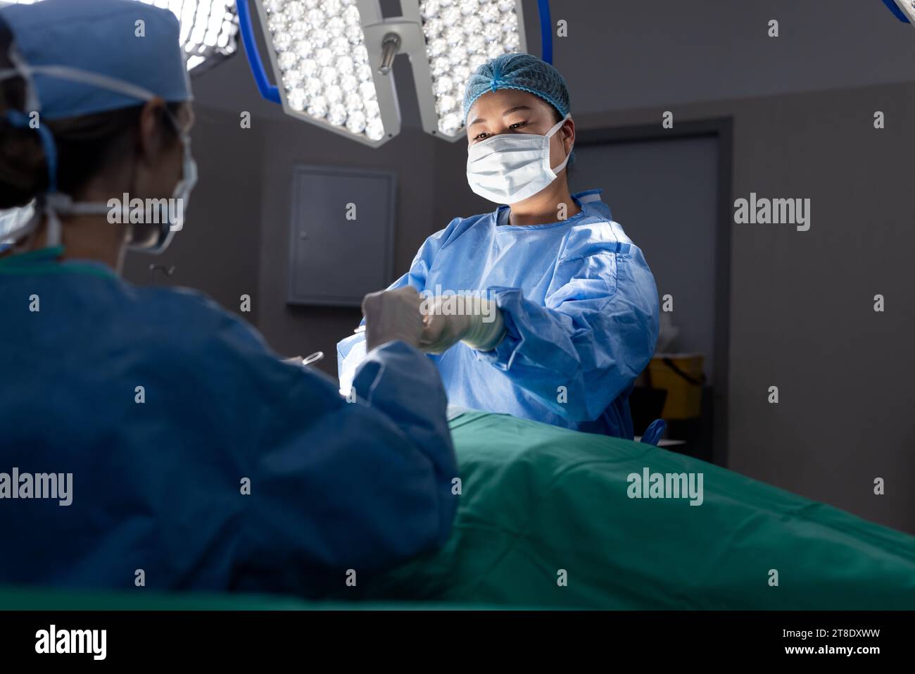 Diverse female doctor with face mask doing surgery in group of doctors ...
