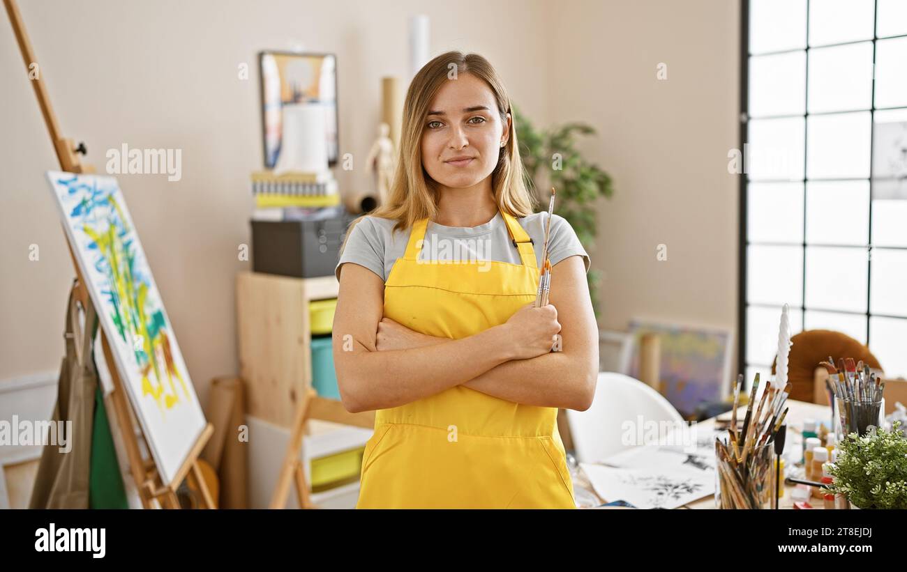 Confident young blonde artist, smiling with arms crossed, standing amid ...
