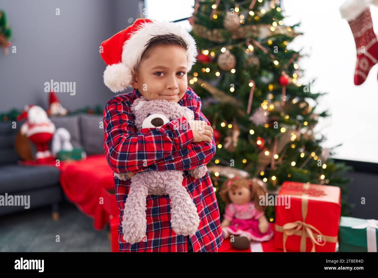 Adorable hispanic boy hugging teddy bear standing by christmas tree at ...