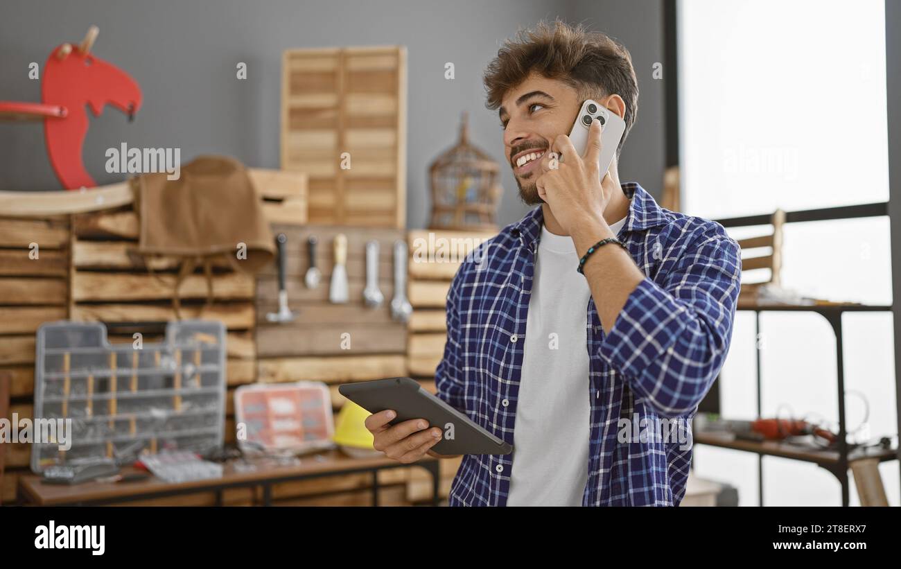 Handsome young arab man carpenter talking on smartphone, beaming with smiling face, engrossed in ...