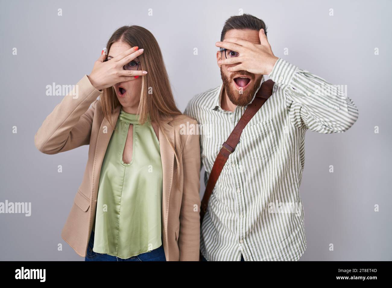 Young couple standing over white background peeking in shock covering ...