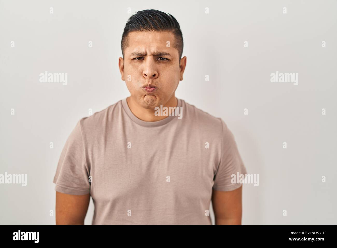 Hispanic young man standing over white background puffing cheeks with ...